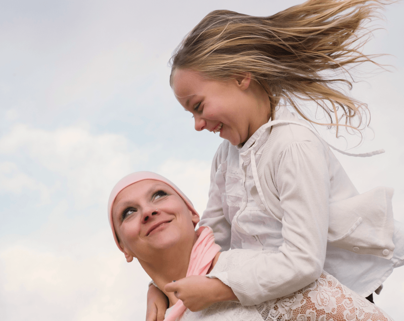 A mother joyfully holding her daughter, both smiling against a cloudy sky.
